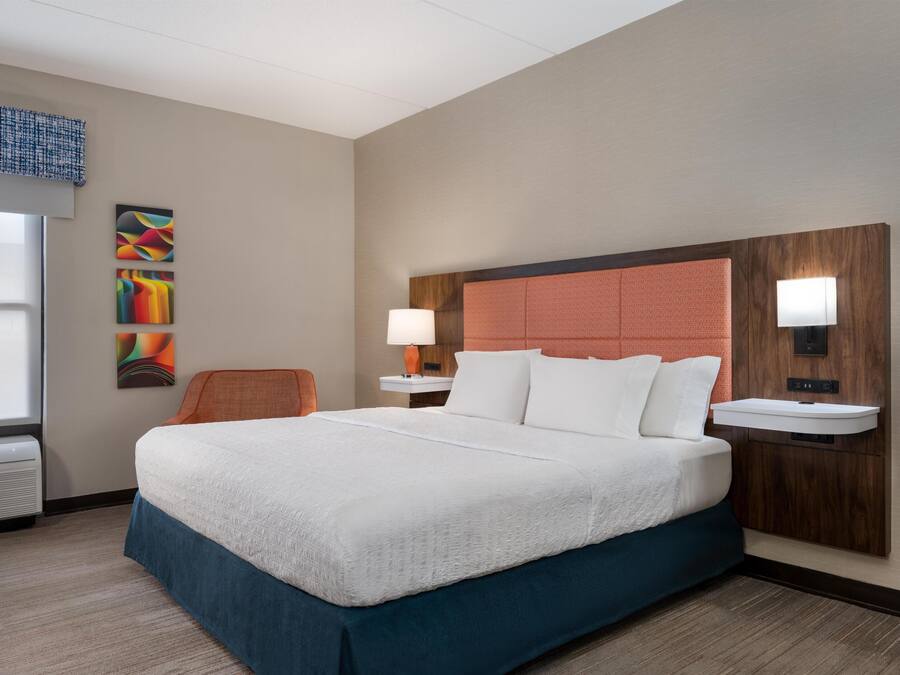 Quarto de hotel com uma cama grande, duas mesas de cabeceira com abajures, uma janela com vista e uma parede com arte abstrata.