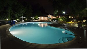 Piscina noturna com espreguiçadeiras e guarda-sóis, cercada por árvores e um edifício iluminado.