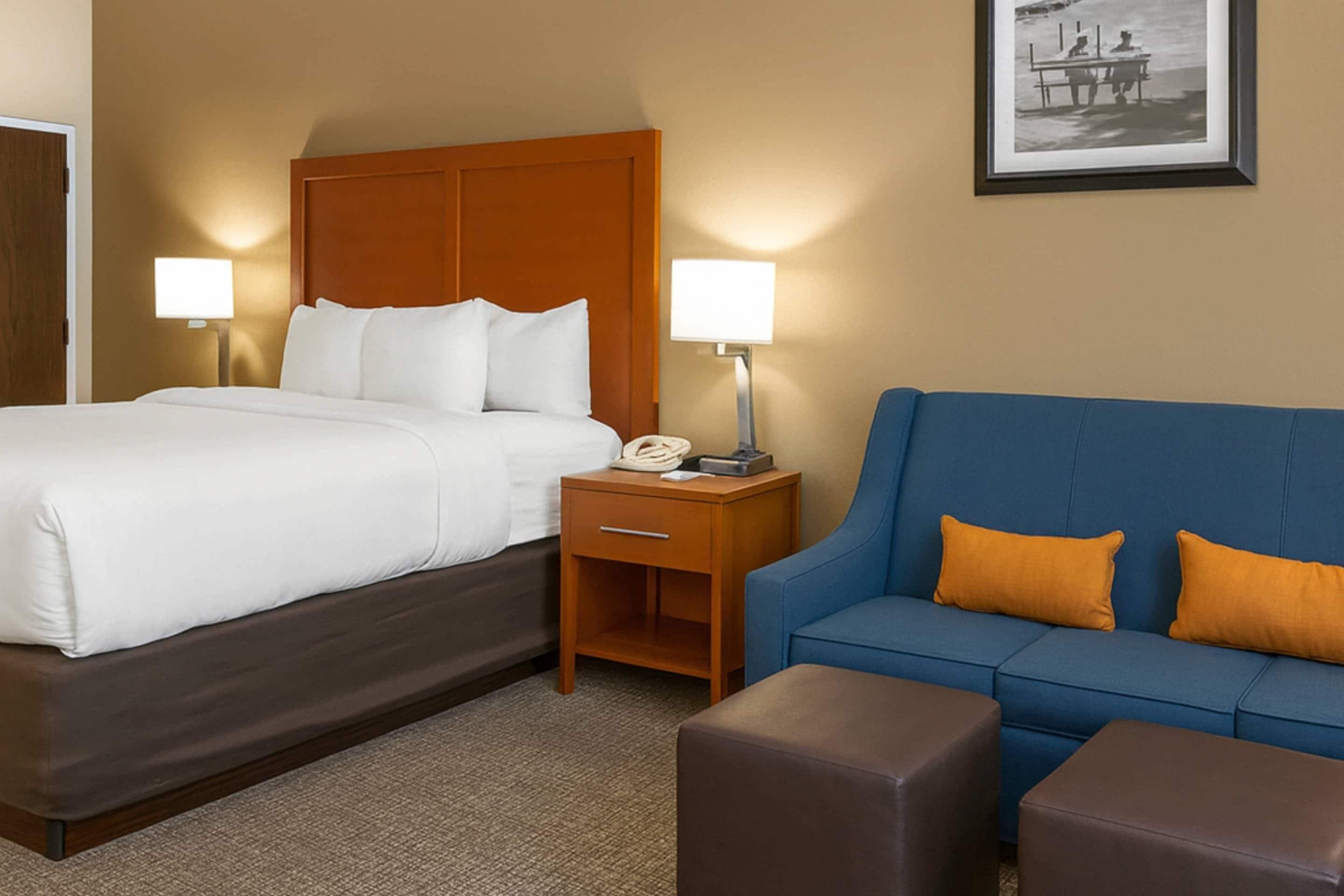 Quarto de hotel com uma cama, mesa de cabeceira com abajur, sofá azul com almofadas laranja e um quadro na parede.