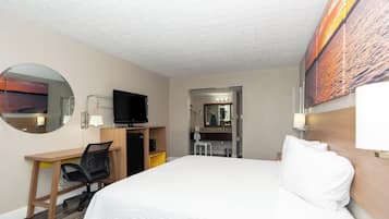 Quarto de hotel com cama, escrivaninha com cadeira, televisão, espelho e banheiro visível através de uma porta aberta.