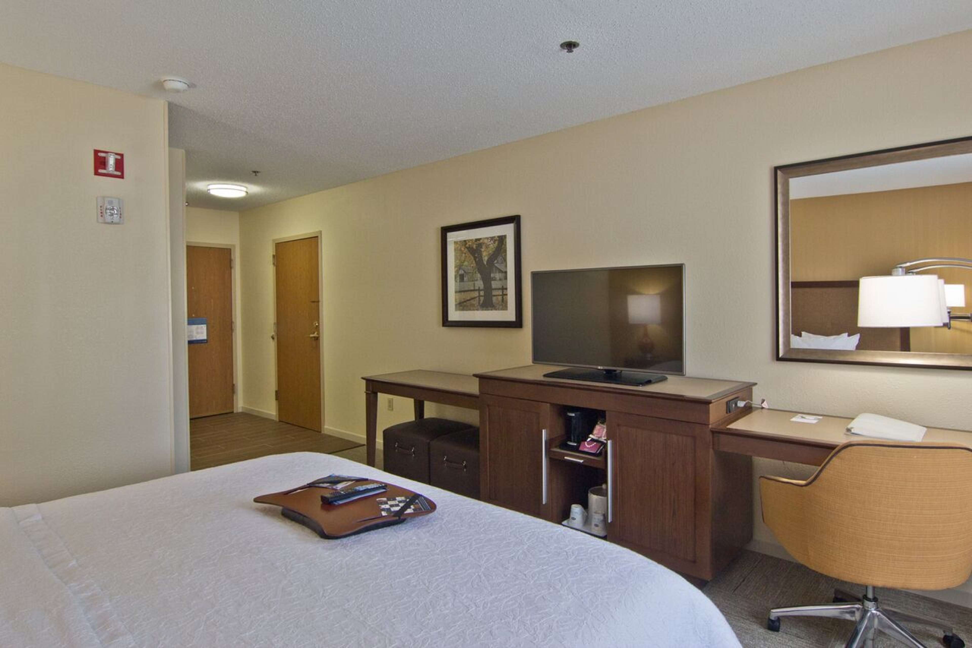 Quarto de hotel com uma cama, escrivaninha, cadeira, televisão e espelho.