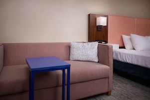 Quarto de hotel com sofá rosa, uma pequena mesa azul, cama com roupa de cama branca e uma luminária fixada na parede.