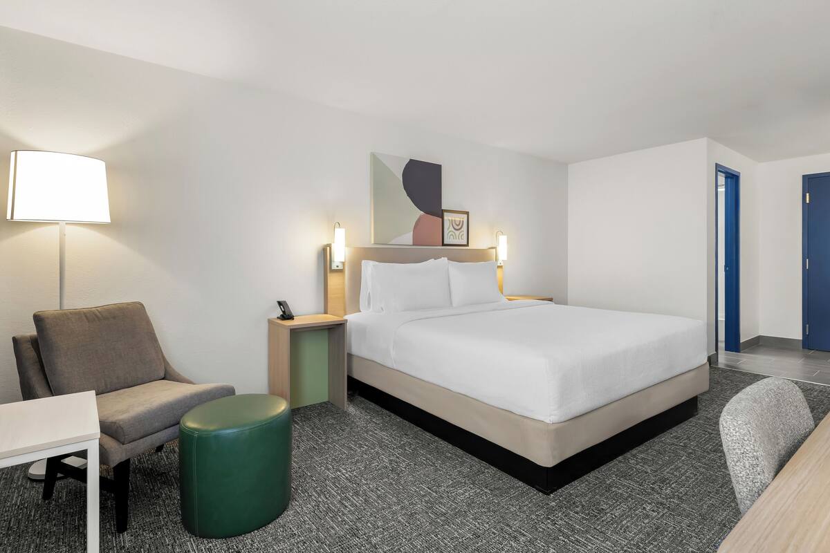 Quarto de hotel com cama, poltrona, mesinha, luminária e um quadro na parede.