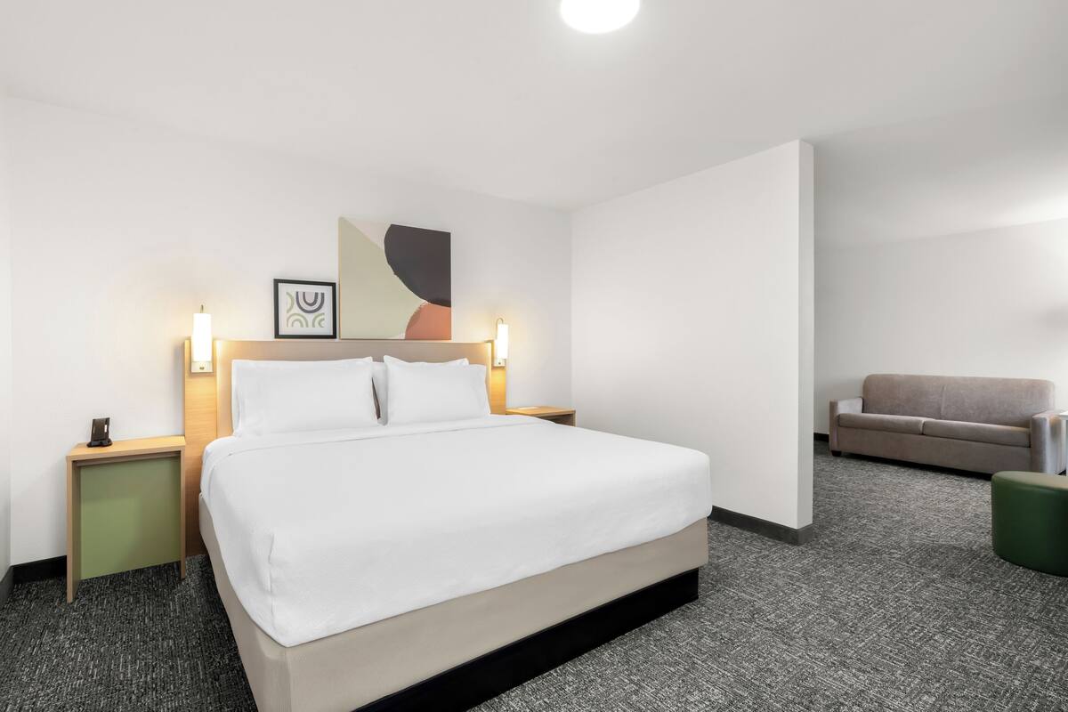 Quarto de hotel com uma cama grande, duas mesas de cabeceira com luminárias, uma mesinha lateral verde, um sofá bege e um banquinho verde.