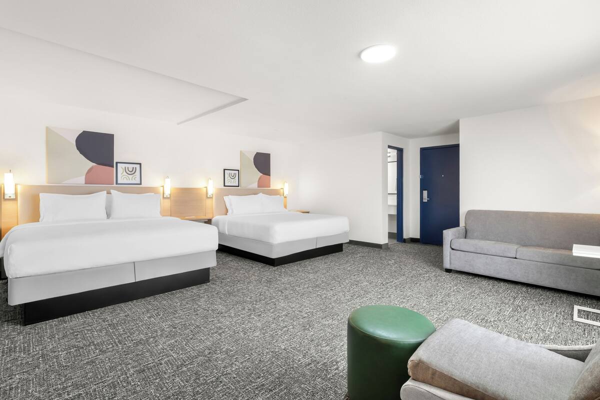 Quarto de hotel com duas camas, um sofá, um banquinho verde e obras de arte nas paredes.