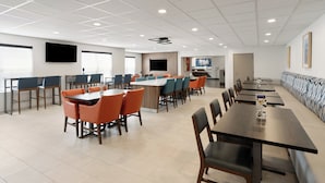 Uma sala de conferências moderna com várias mesas e cadeiras, uma tela grande e uma área de estar com um banco estampado.