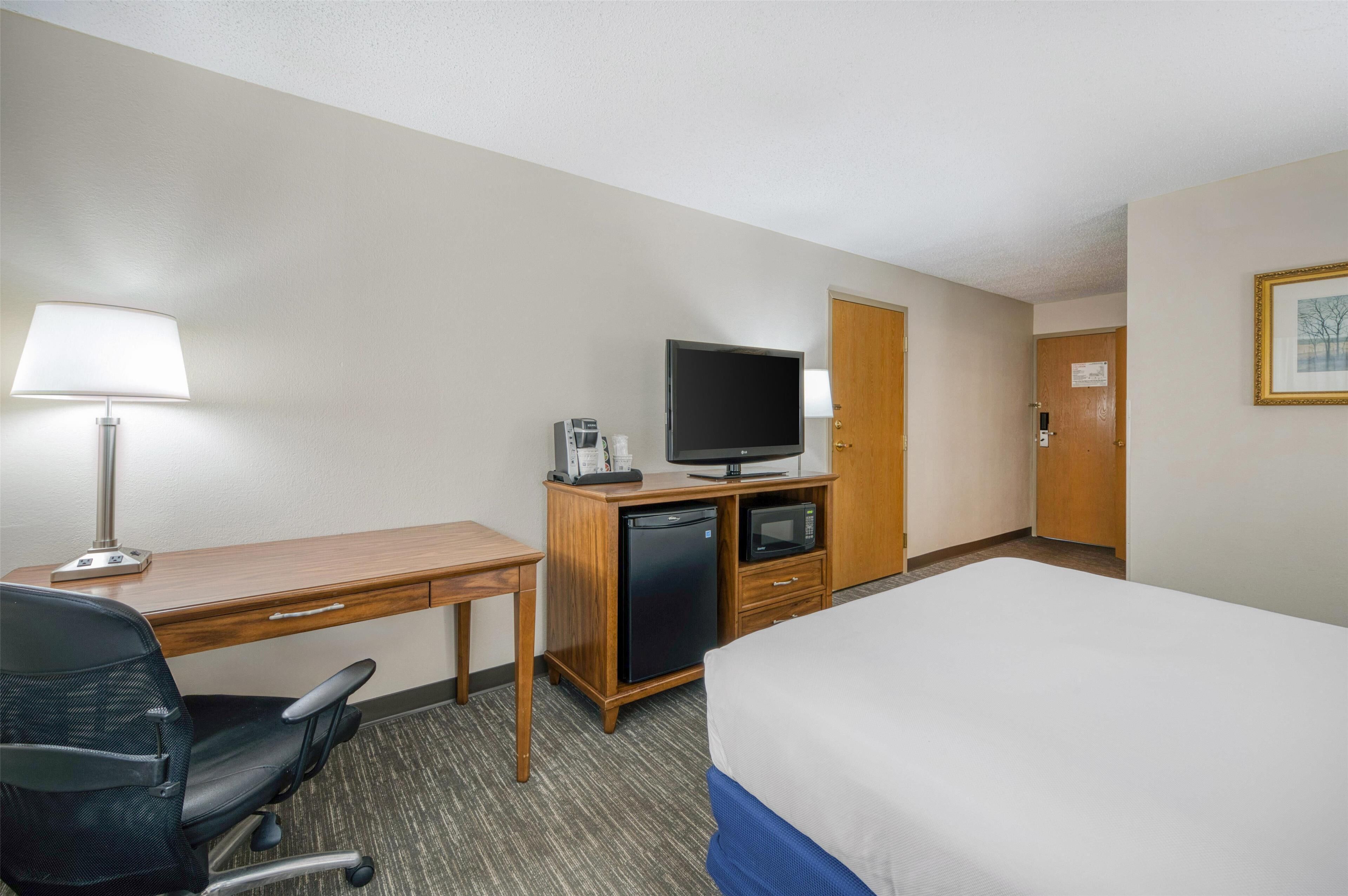 Quarto de hotel com cama, escrivaninha, cadeira, televisão, frigorífico e micro-ondas.
