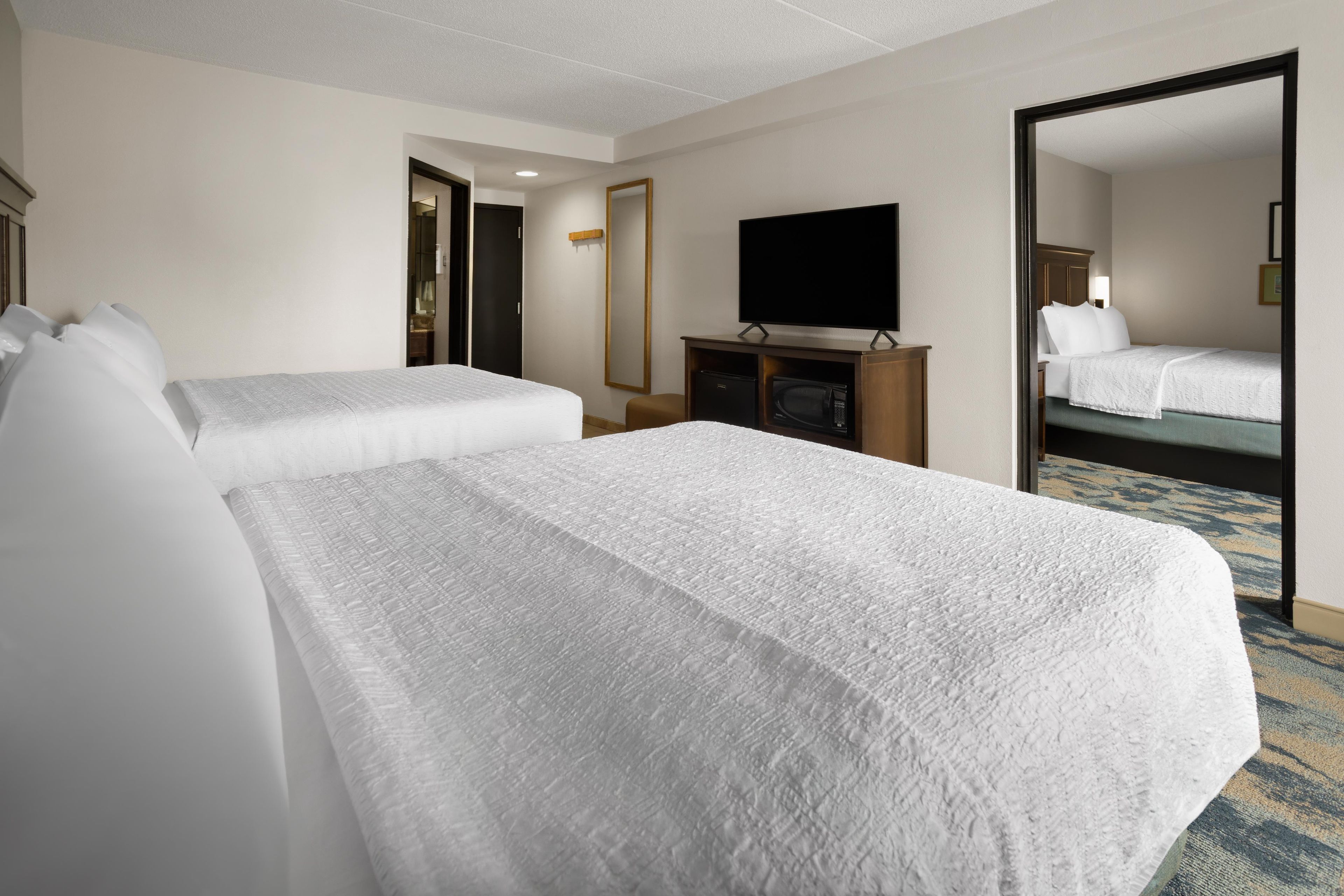 Quarto de hotel com duas camas, televisão, espelho e porta que dá para outro cômodo.