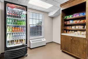 Máquina automática com diversas bebidas e lanches, uma janela com persianas e um armário de madeira com petiscos.