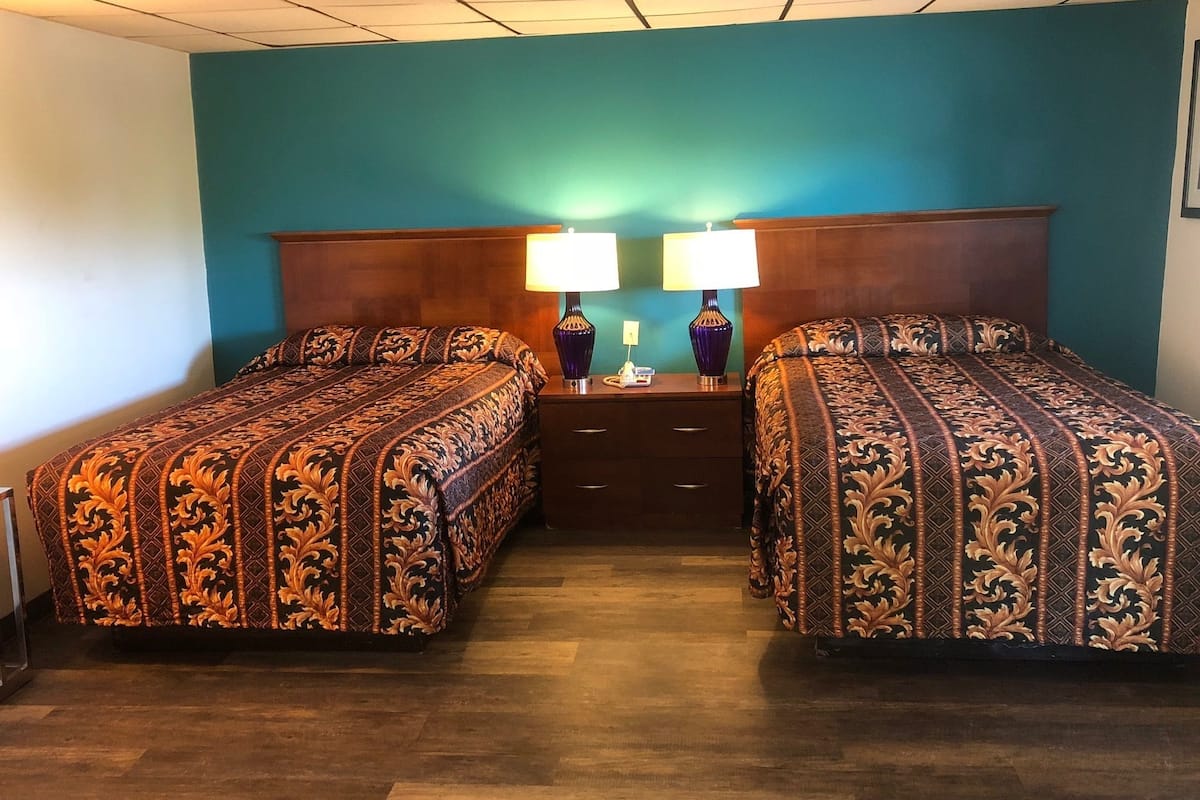 Quarto de hotel com duas camas, cada uma com colcha estampada, cabeceira de madeira e mesa de cabeceira com abajur entre as camas.