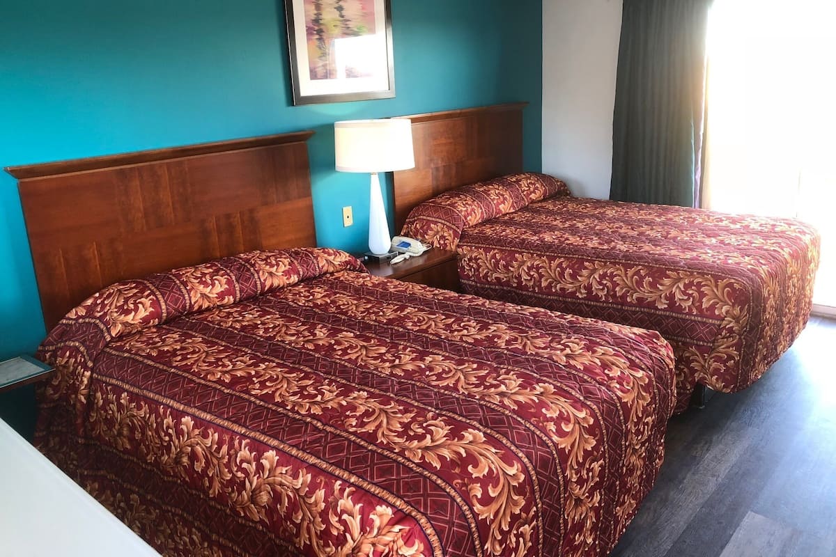 Quarto de hotel com duas camas, cabeceira de madeira, mesa de cabeceira com abajur e um quadro na parede.