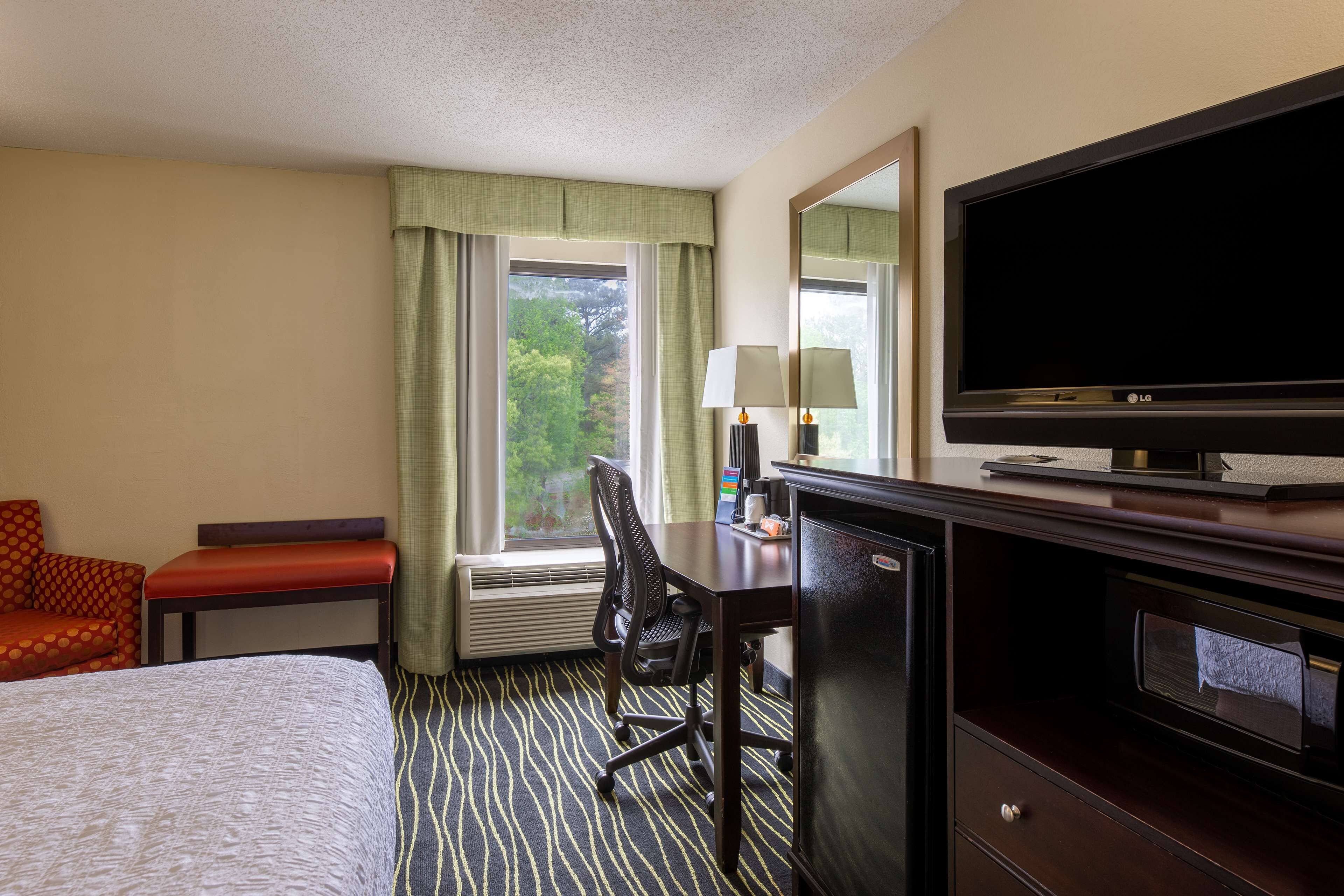 Quarto de hotel com cama, escrivaninha com televisão, cadeira, abajur e janela com cortinas.
