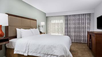 Quarto de hotel com uma cama grande, uma mesa de cabeceira com abajur, televisão e janela com cortinas estampadas.