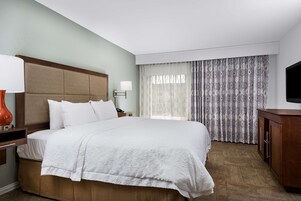 Quarto de hotel com uma cama grande, uma mesa de cabeceira com abajur, televisão e janela com cortinas estampadas.