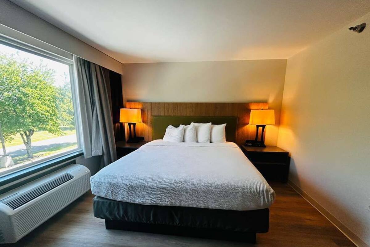 Quarto de hotel com uma cama grande, duas luminárias de cabeceira, uma janela com cortinas e um aquecedor fixado na parede.