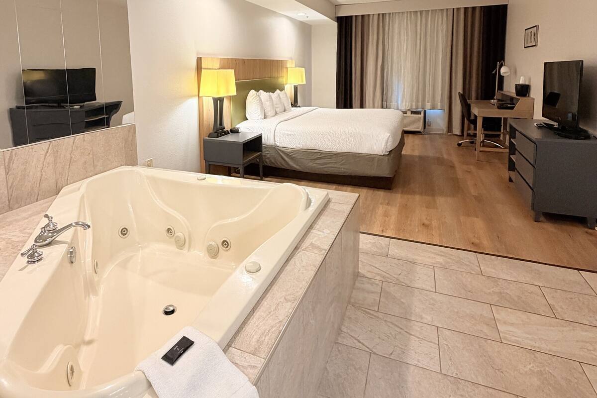 Quarto de hotel moderno com uma banheira grande, cama com cabeceira de madeira, escrivaninha com cadeira e televisão.