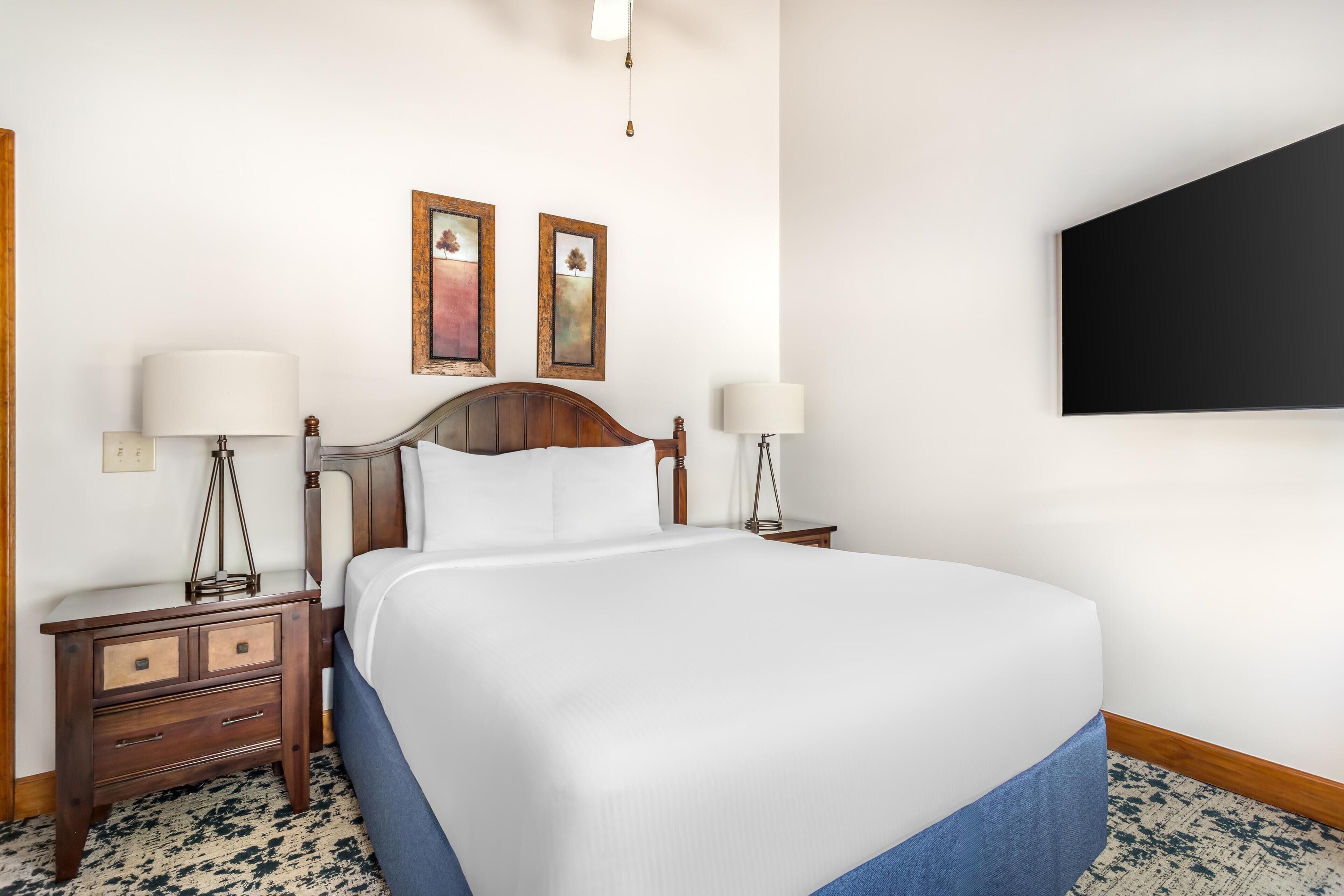 Quarto de hotel com cama grande, duas mesinhas de cabeceira, televisão de tela plana na parede e duas fotos emolduradas acima da cama.