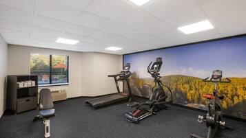 Uma sala de fitness com esteira, elíptica e bicicleta ergométrica, uma TV fixada na parede e uma janela com persianas.