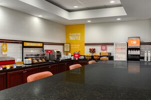 Espaço de café da manhã com balcão, assentos e diferentes estações de serviço de alimentos, incluindo estação de waffles e dispensador de sucos.