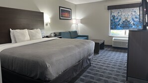 Quarto de hotel com uma cama grande, uma escrivaninha, uma cadeira, uma televisão e uma janela com padrão geométrico.