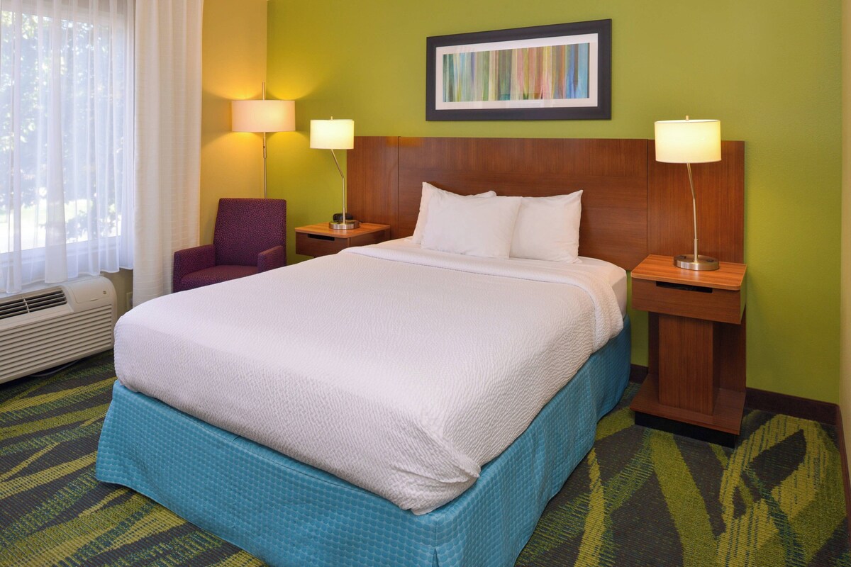Quarto de hotel com uma cama grande, duas mesas de cabeceira com luminárias, uma cadeira e uma janela com cortinas.