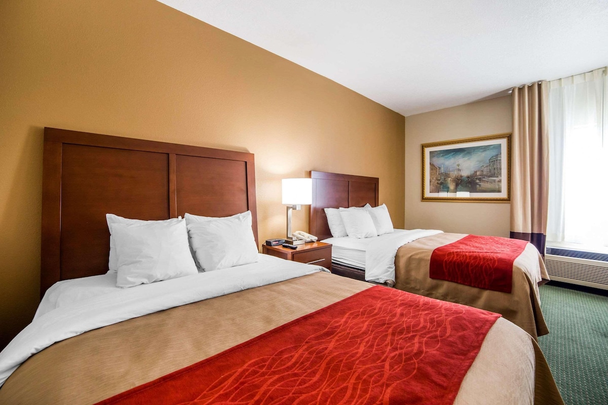 Quarto de hotel com duas camas, paredes bege, um quadro na parede e uma janela com cortinas.