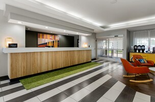 Uma área de recepção de hotel com um balcão de madeira, uma pintura abstrata colorida, um tapete de grama artificial verde e cadeiras laranjas.