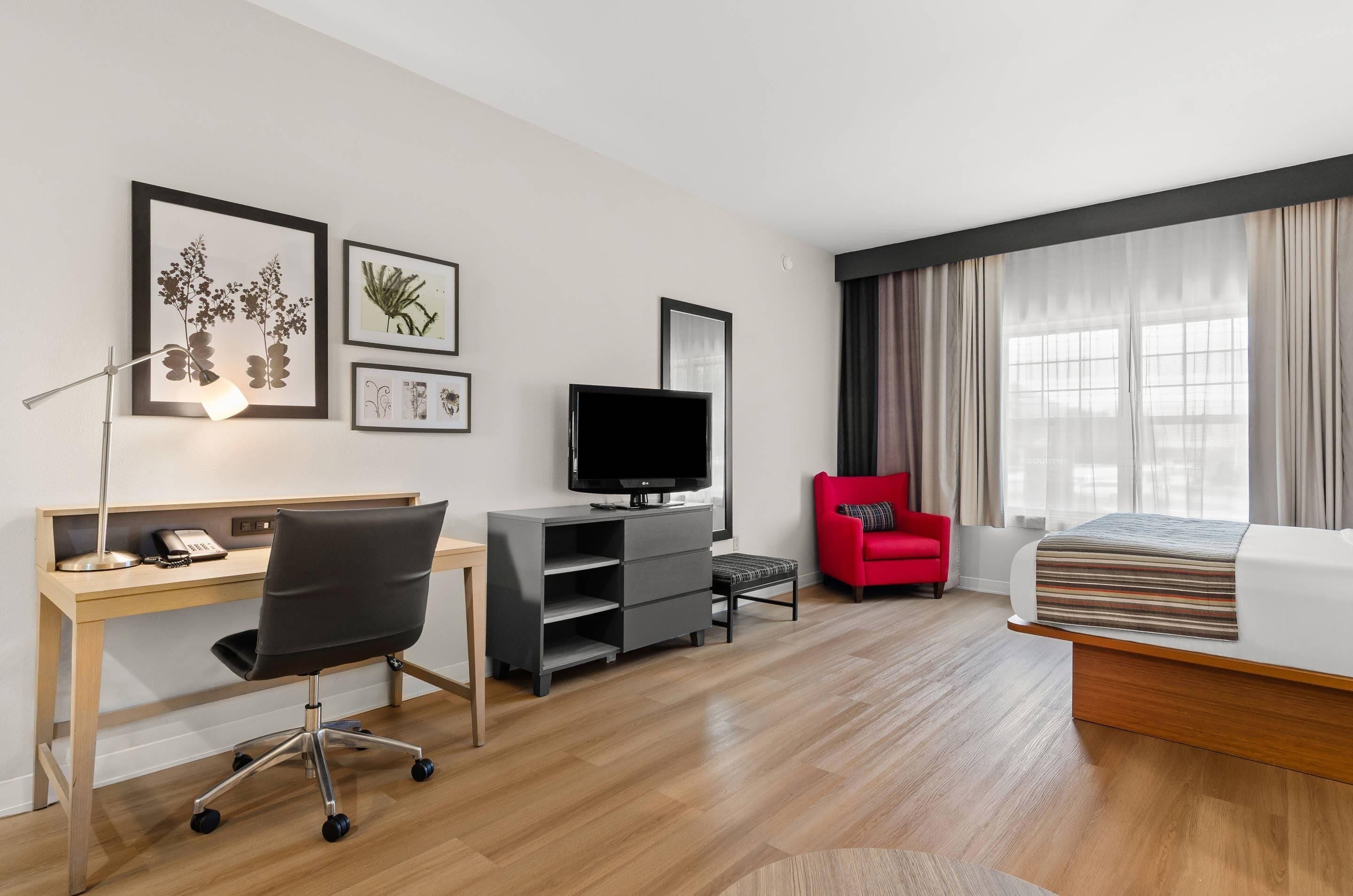 Quarto de hotel moderno com escrivaninha de madeira, televisão, poltrona vermelha e cama com manta listrada.