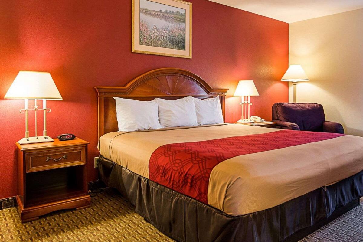 Quarto de hotel com uma cama grande, duas mesas de cabeceira com abajures, uma cadeira e um quadro na parede.