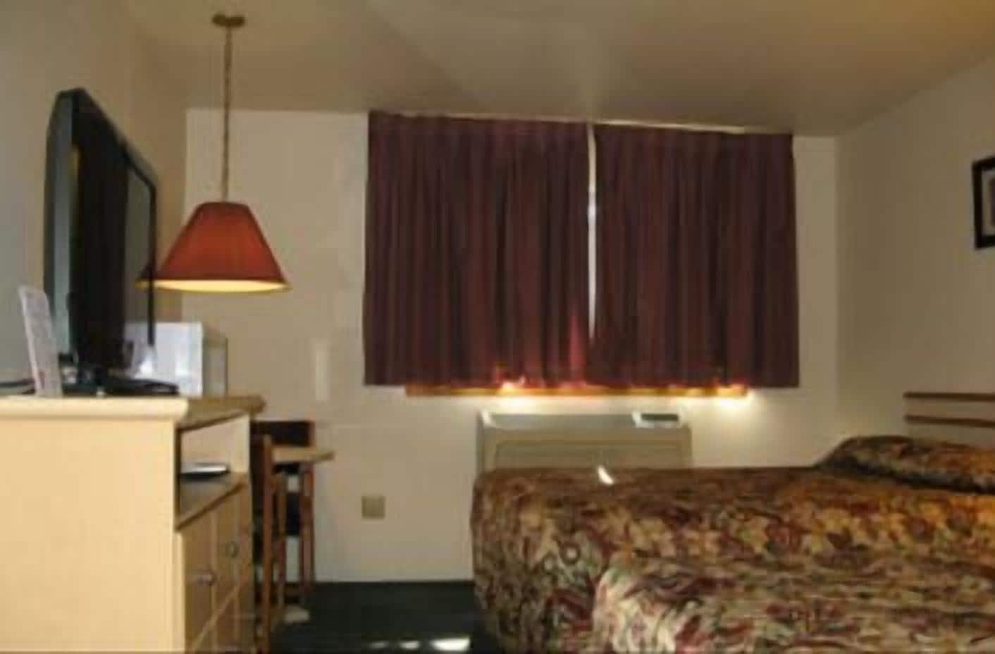 Um quarto pequeno e modesto, com uma cama, uma escrivaninha, uma cadeira e uma televisão.