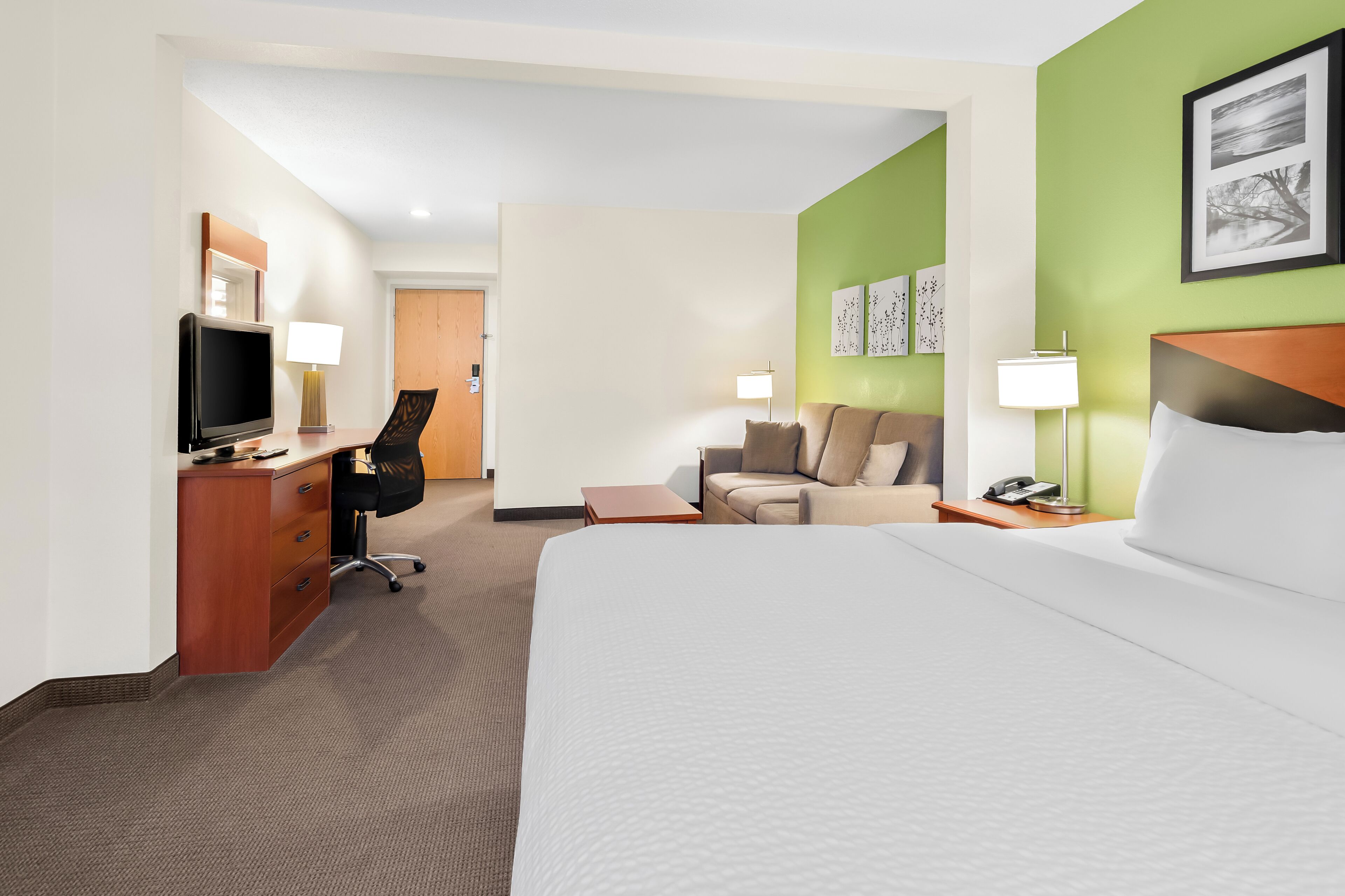 Quarto de hotel com uma cama grande, uma escrivaninha com computador, uma cadeira, um sofá e uma televisão.