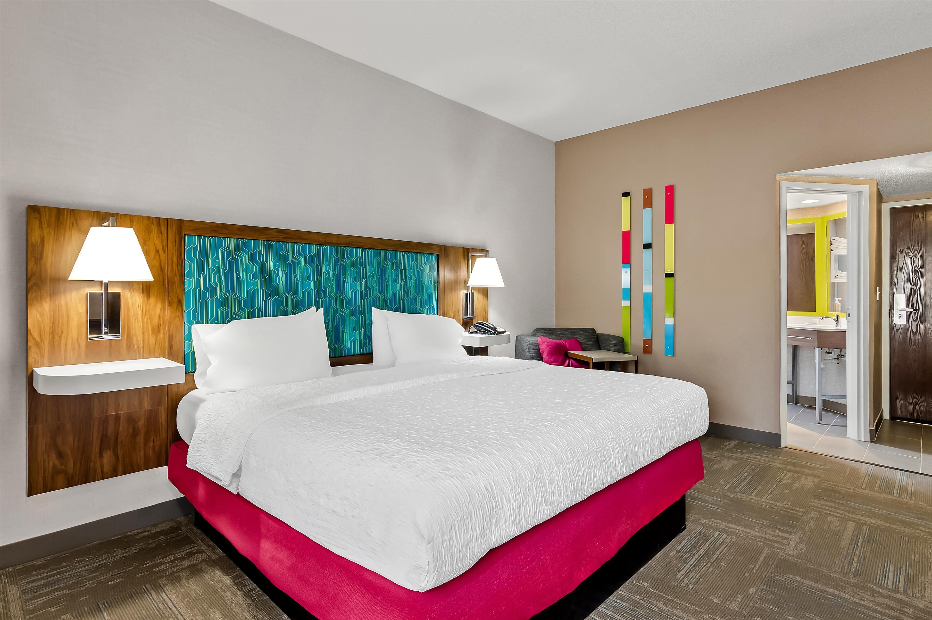 Quarto de hotel com uma cama grande, duas luminárias de cabeceira, uma cabeceira de madeira e uma obra de arte abstrata colorida na parede.