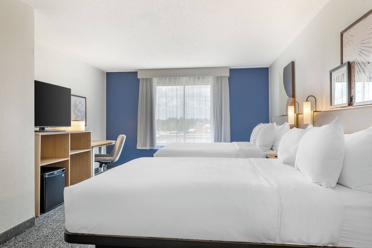 Quarto de hotel com duas camas, uma escrivaninha, uma cadeira, uma televisão e uma janela com cortinas.