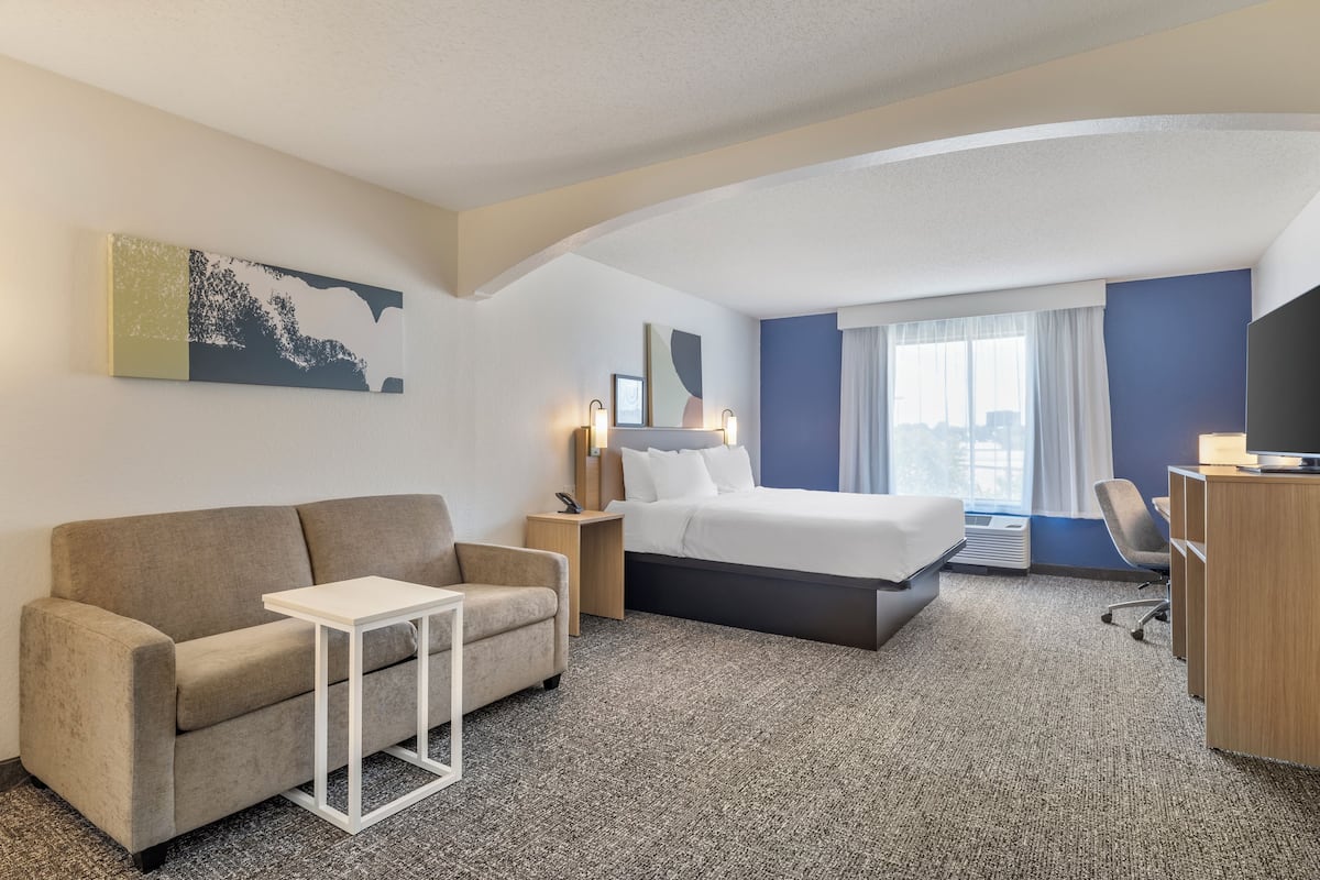Quarto de hotel com cama, sofá, pequena mesa, escrivaninha e televisão.