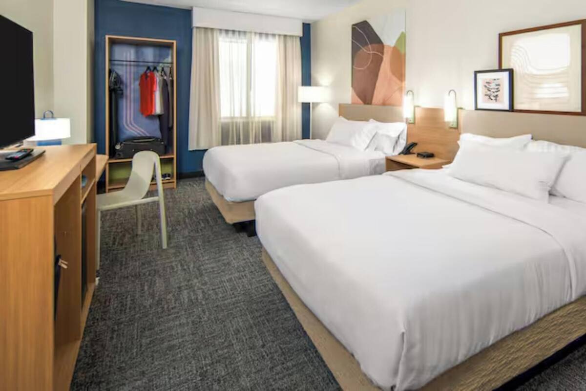 Quarto de hotel com duas camas, uma escrivaninha, uma cadeira, um guarda-roupa e uma janela com cortinas.