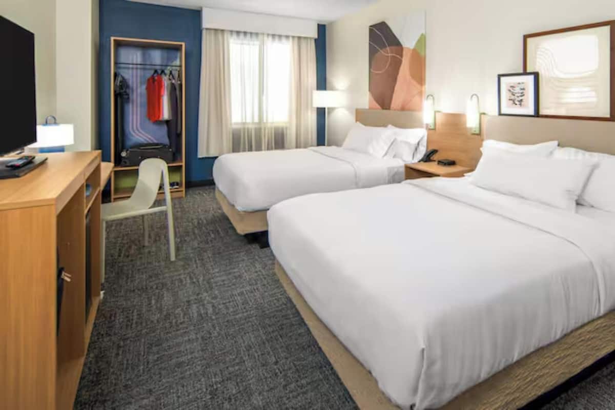Quarto de hotel com duas camas, uma escrivaninha, uma cadeira, um guarda-roupa e uma janela com cortinas.