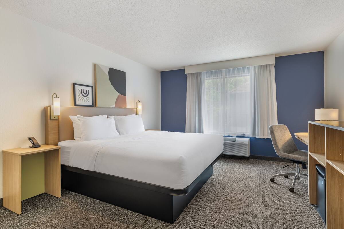 Quarto de hotel com cama grande, mesas de cabeceira, escrivaninha, cadeira e janela com cortinas.