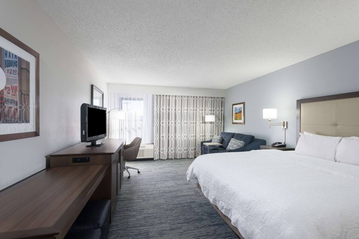 Quarto de hotel com cama, escrivaninha com televisão, cadeira, luminária e janela com cortinas.