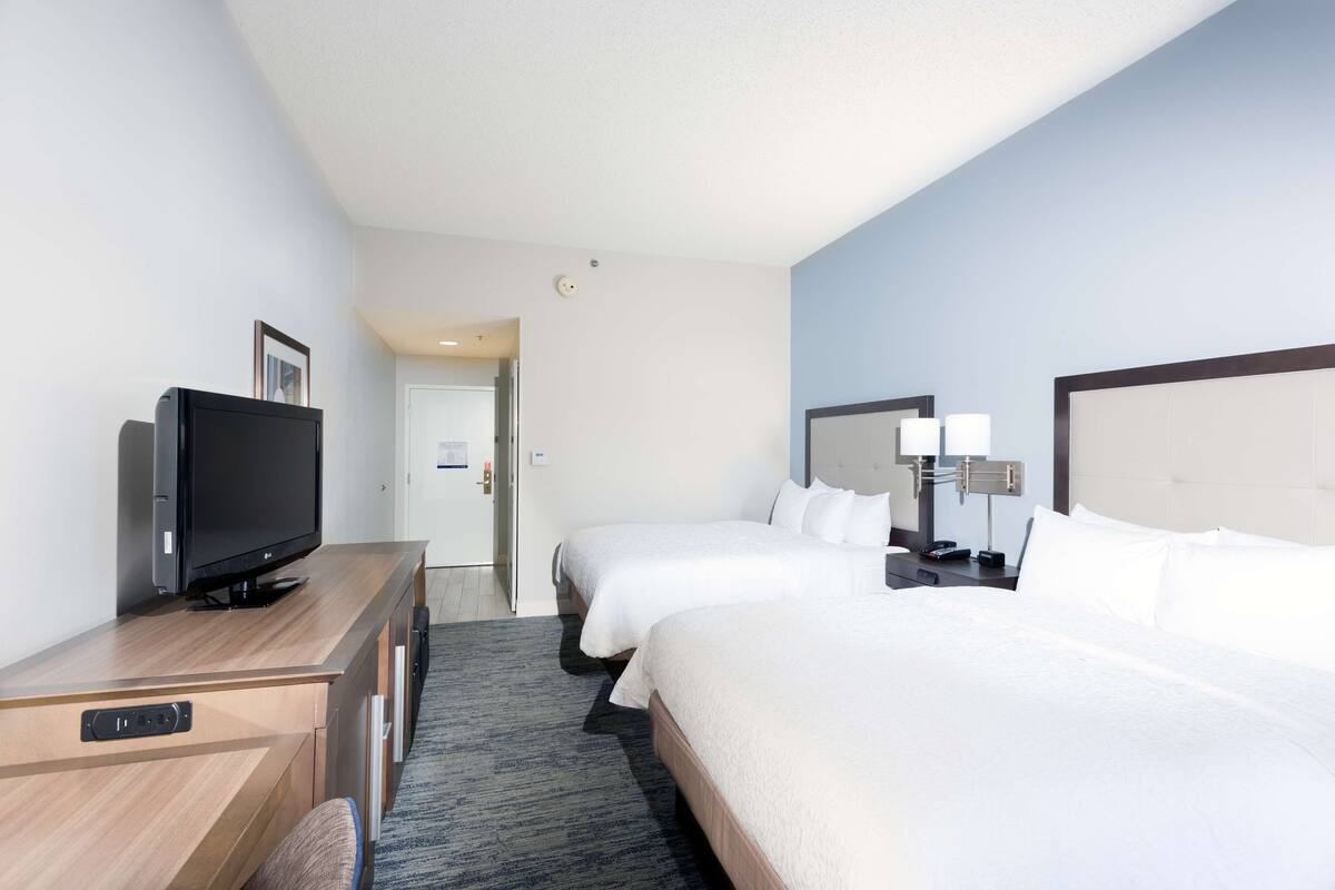 Quarto de hotel com duas camas, uma televisão, uma mesa e uma luminária.
