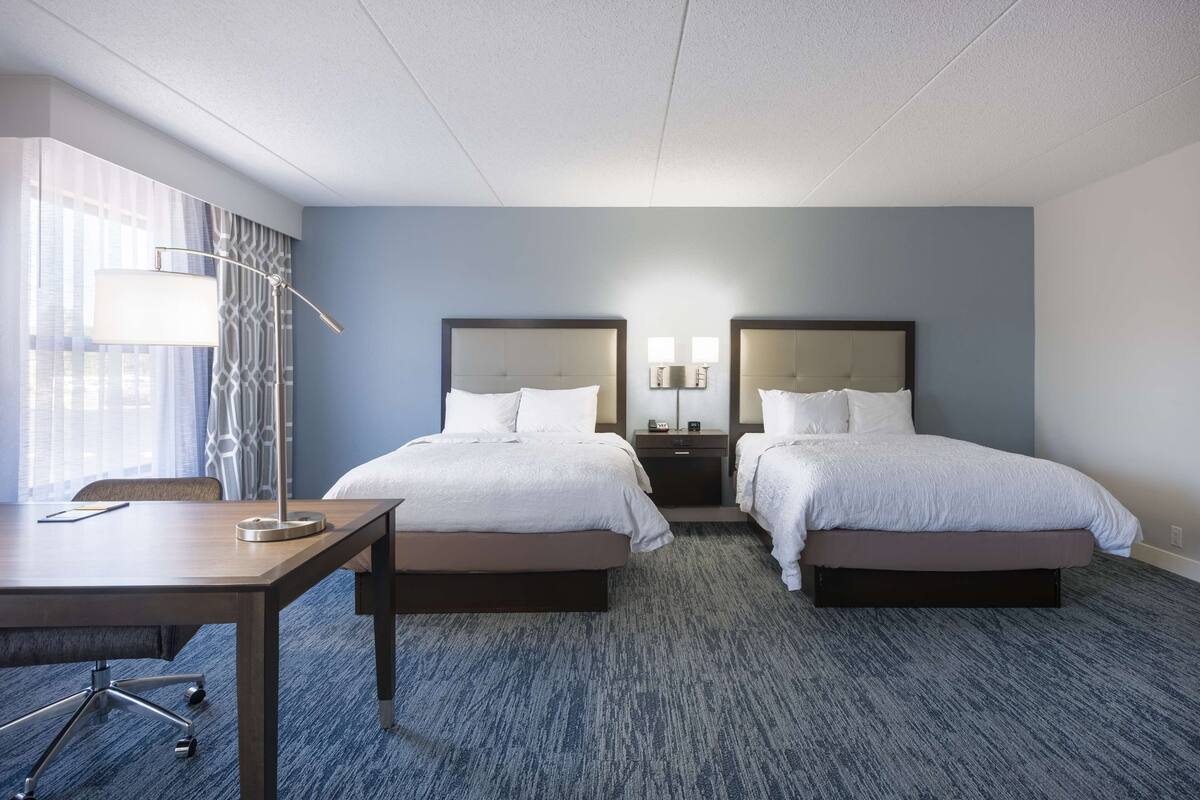 Quarto de hotel com duas camas, uma escrivaninha e uma cadeira. Há uma janela com cortinas e uma luminária na escrivaninha.