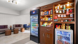 Um espaço de recepção com assentos, um refrigerador Pepsi, prateleiras com lanches e um mini-refrigerador.