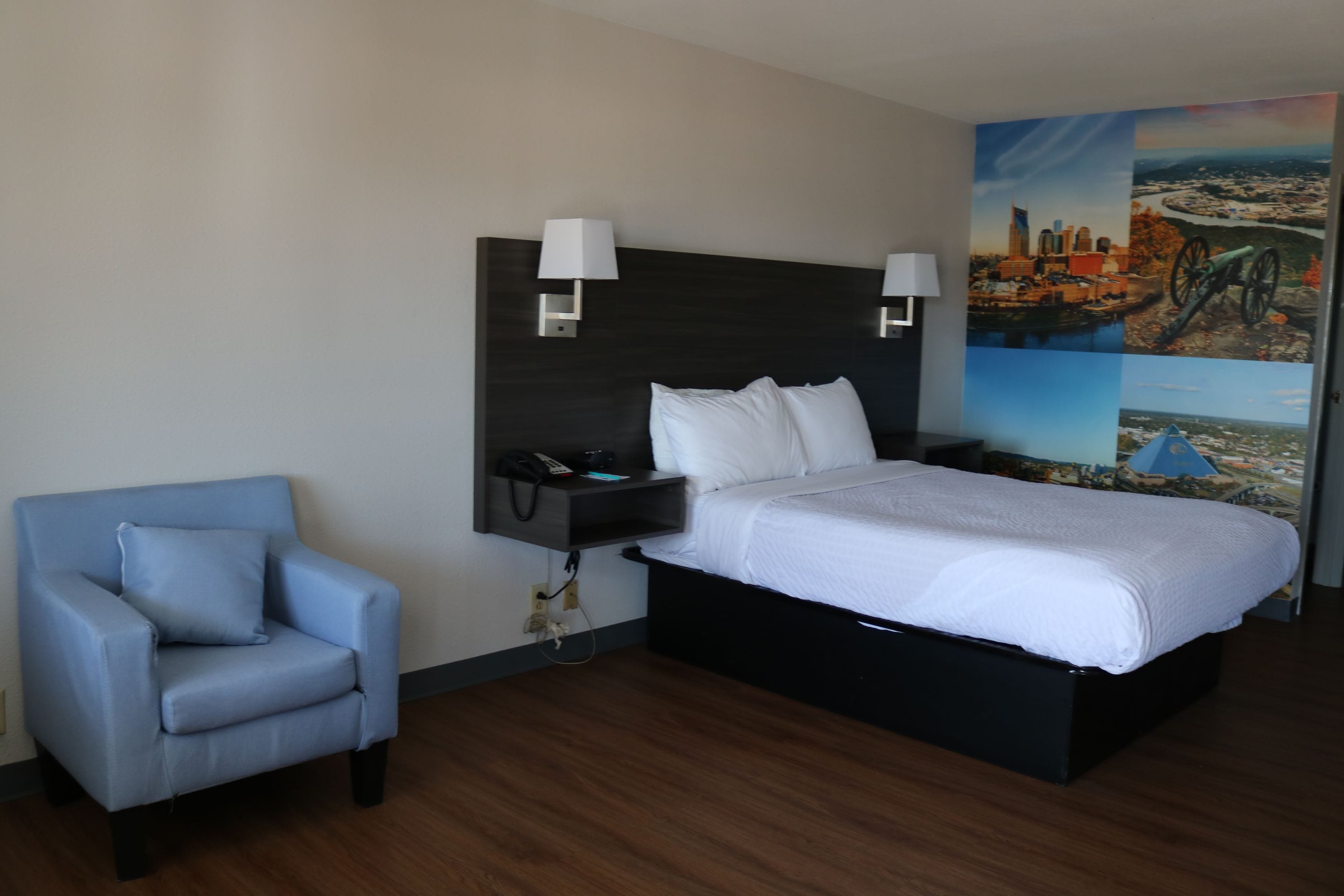 Quarto de hotel com cama, uma cadeira, mesa de cabeceira com telefone e parede com um mural de paisagem urbana.