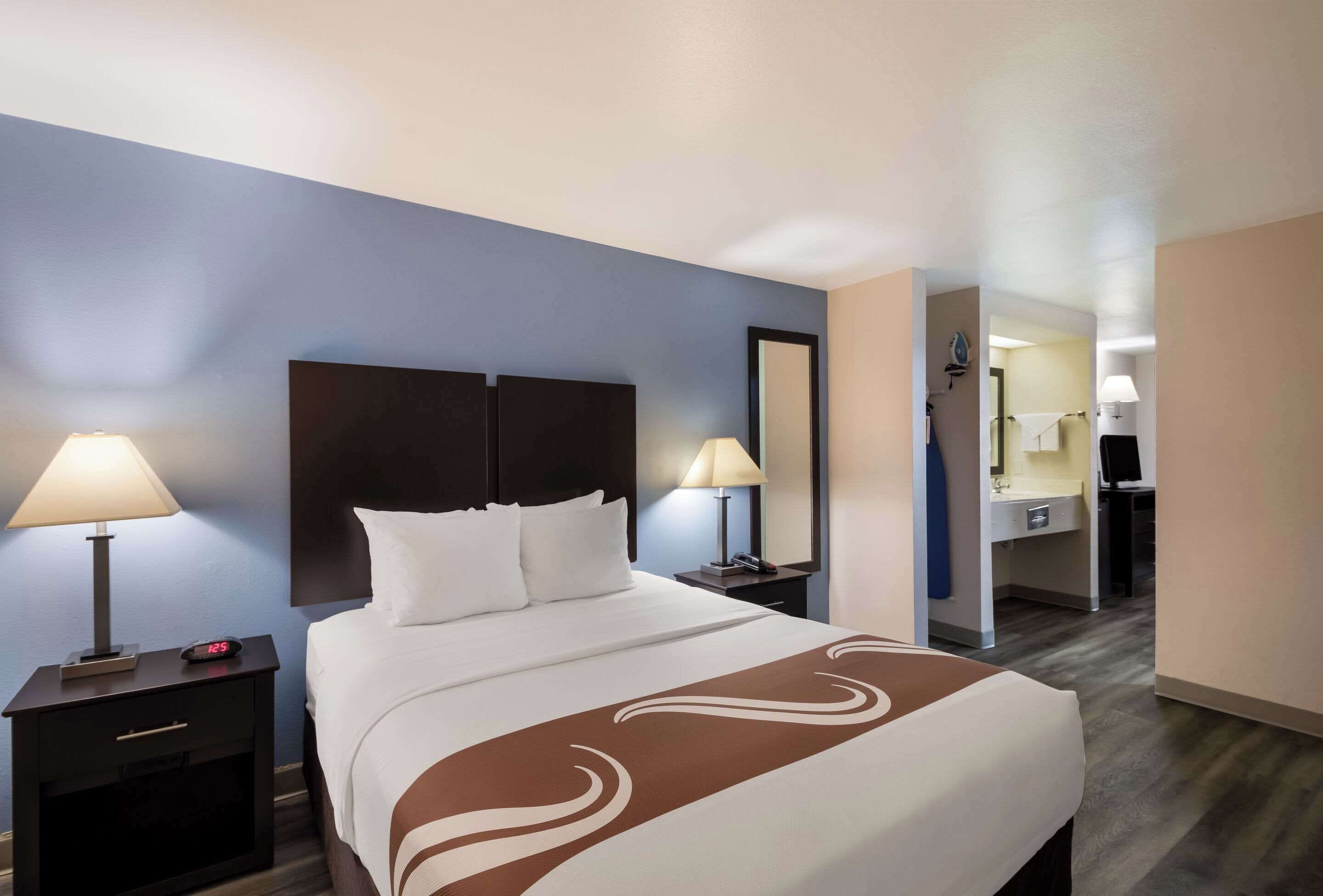 Quarto de hotel com cama, mesas de cabeceira, luminárias, um espelho e um banheiro visível por uma porta aberta.