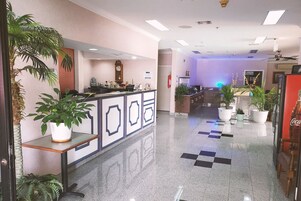Um saguão de hotel com balcão de recepção, plantas em vasos, um relógio e um balcão de check-in.
