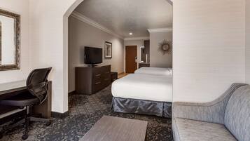 Quarto de hotel com cama, escrivaninha com cadeira, cômoda, televisão e um objeto decorativo na parede.