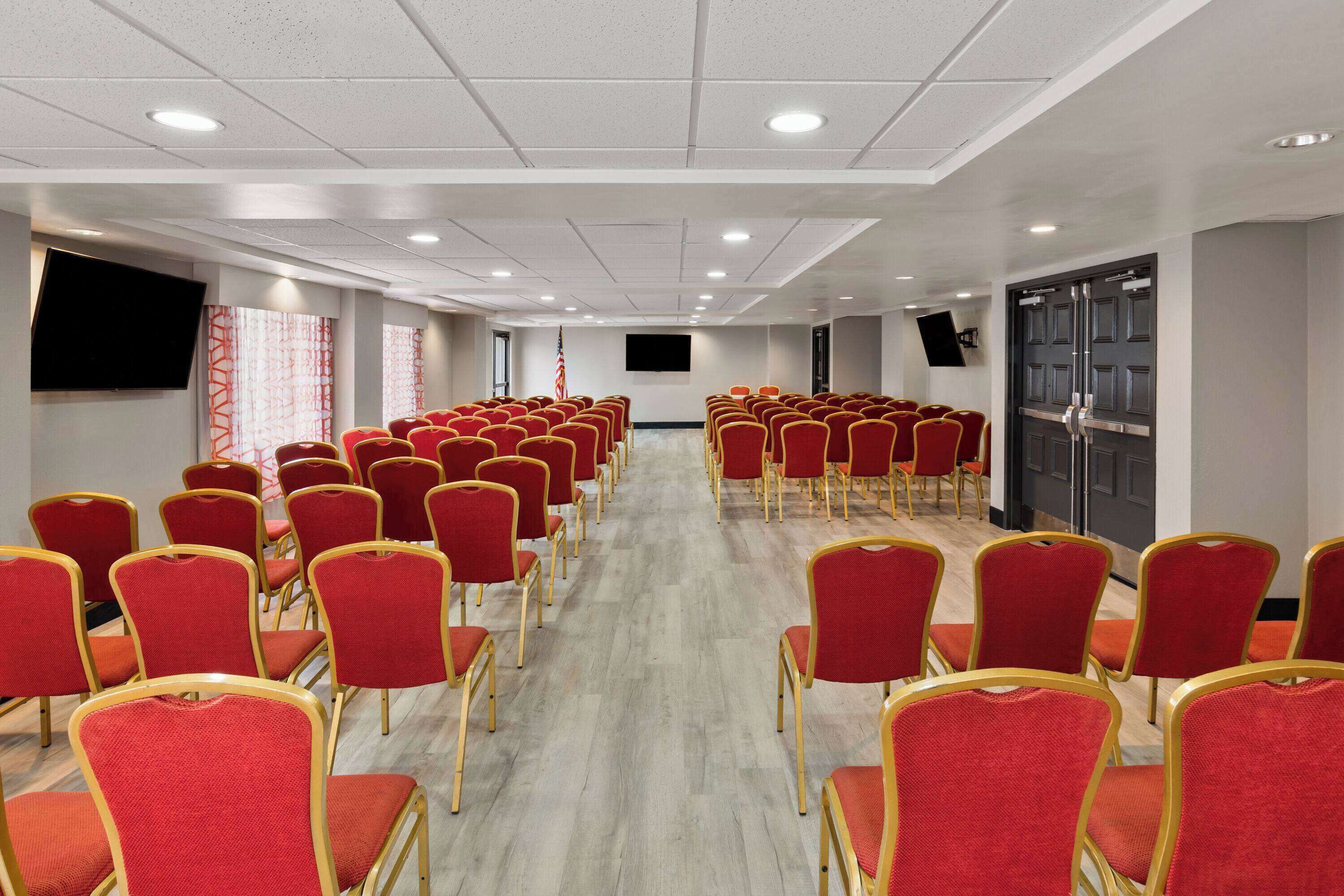 Uma sala de conferência com fileiras de cadeiras vermelhas e uma tela grande na parede.