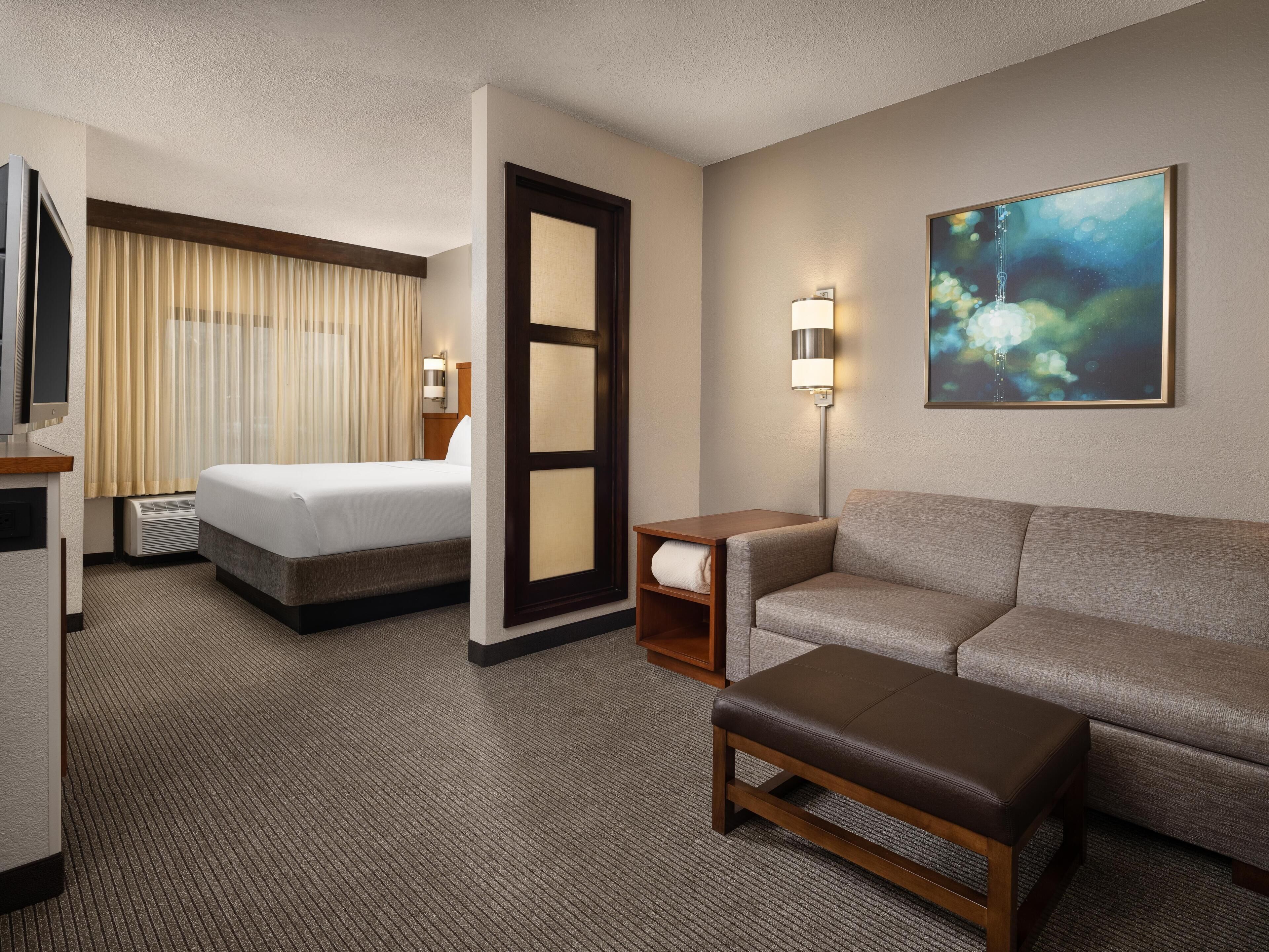 Quarto de hotel com cama, sofá, mesinha, luminária e um quadro na parede.