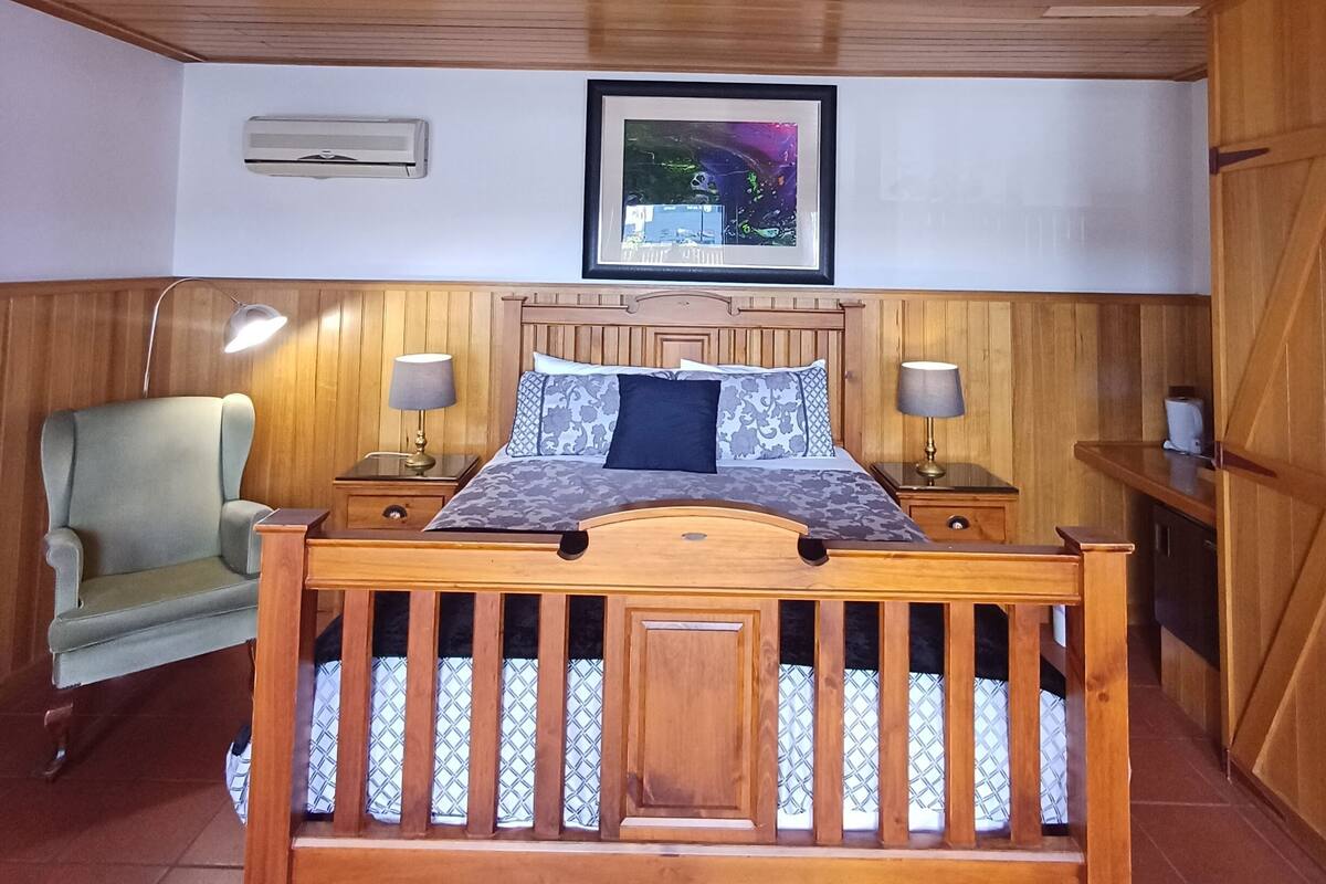 Uma cama de madeira com cabeceira, duas mesas de cabeceira com abajures, uma cadeira e um quadro pendurado na parede.
