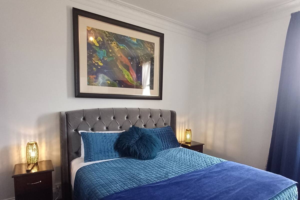 Cama bem arrumada com colcha azul, cabeceira estofada e uma pintura abstrata emoldurada acima.