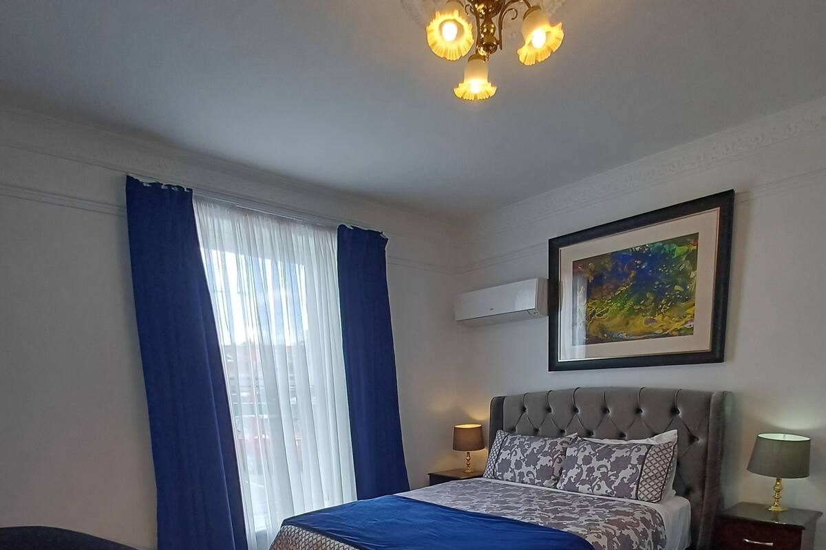 Quarto de hotel com uma cama, uma escrivaninha, uma cadeira, um abajur e um lustre.